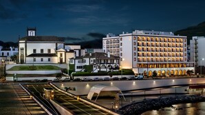 Um hotel com vários andares, uma entrada destacada com área coberta e um estacionamento com vários carros.
