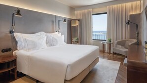 Quarto de hotel moderno com uma cama grande, duas mesas de cabeceira, uma escrivaninha, uma cadeira e vista para o mar.