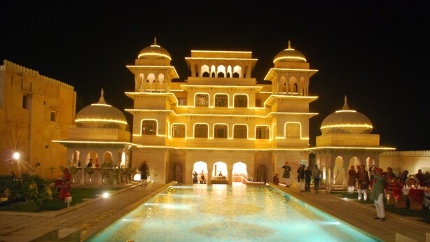 Um edifício grande e iluminado com várias cúpulas e arcos, uma piscina central e pessoas reunidas ao redor.