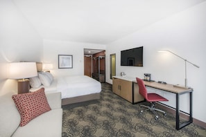 Quarto de hotel com cama, escrivaninha, cadeira, abajur, televisão e carpete estampado.