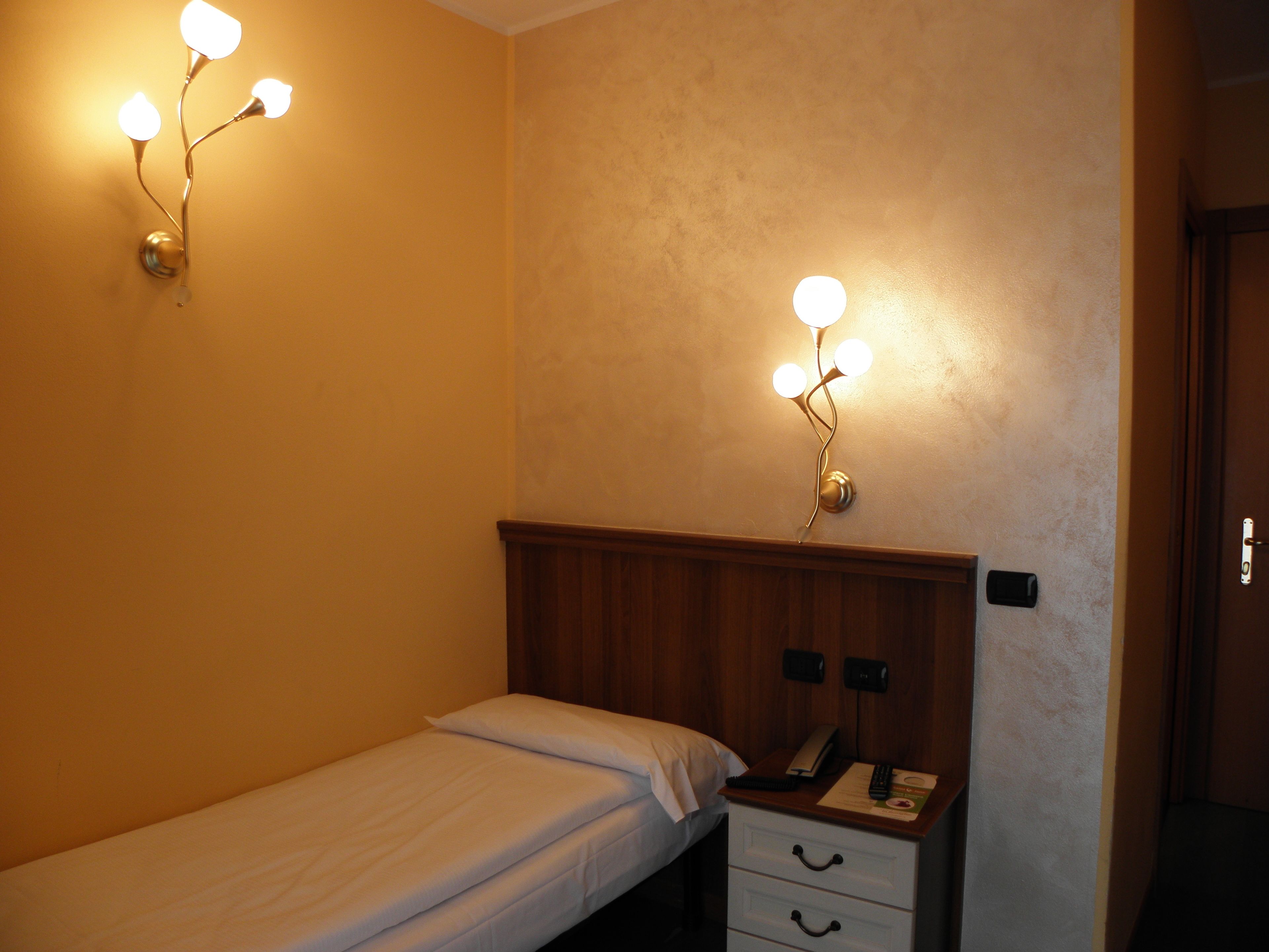 Quarto pequeno com uma cama de solteiro, cabeceira de madeira, mesa de cabeceira com telefone e controle remoto, e luminárias fixadas na parede.