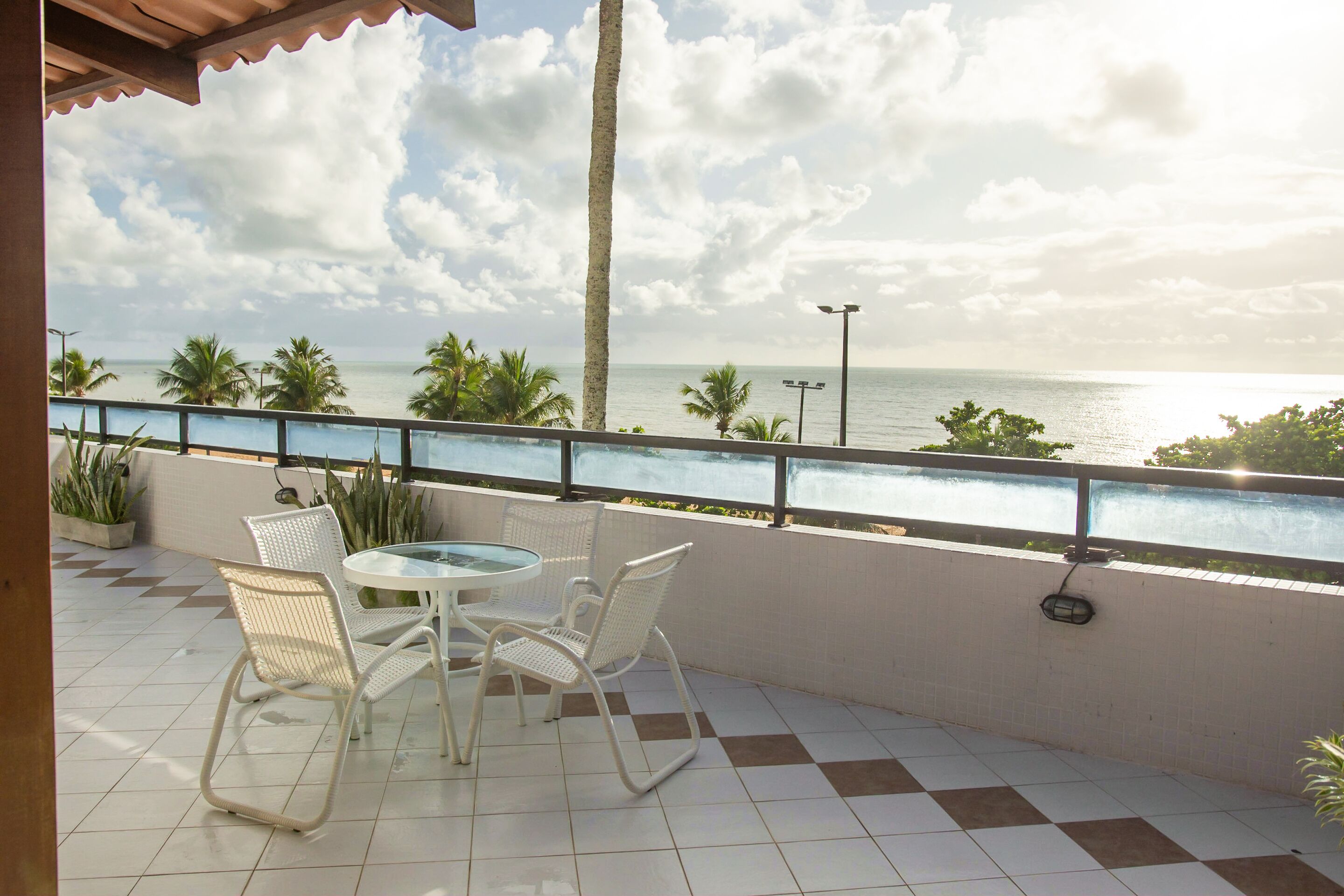Uma varanda com mesa e cadeiras de vidro, com vista para uma praia com palmeiras e céu claro.