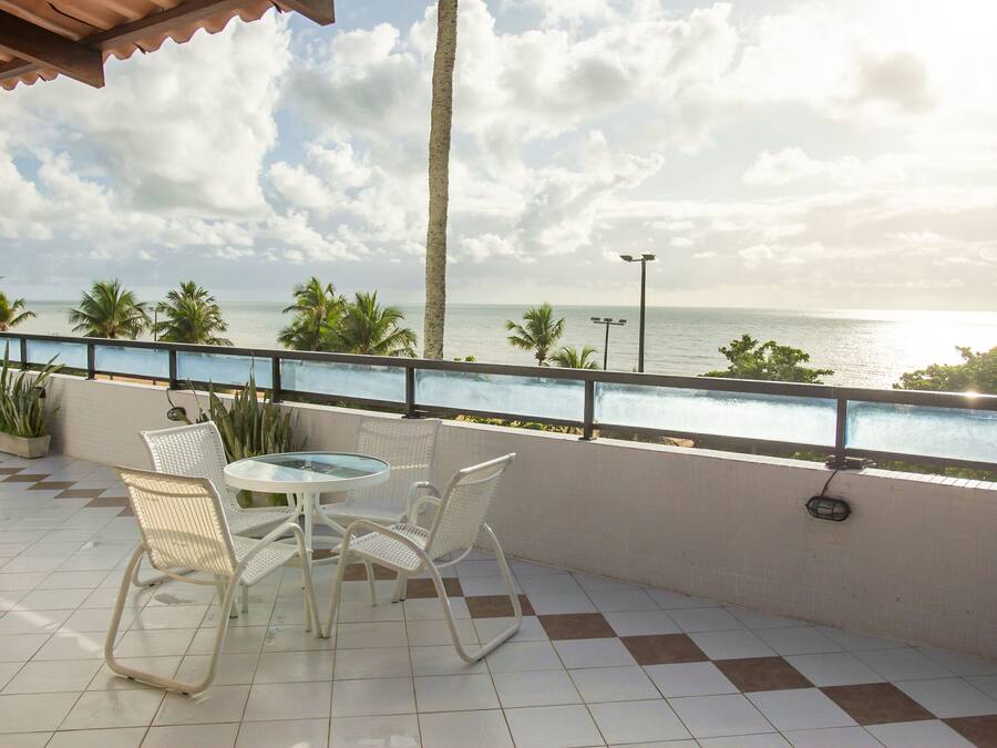 Uma varanda com mesa e cadeiras de vidro, com vista para uma praia com palmeiras e céu claro.