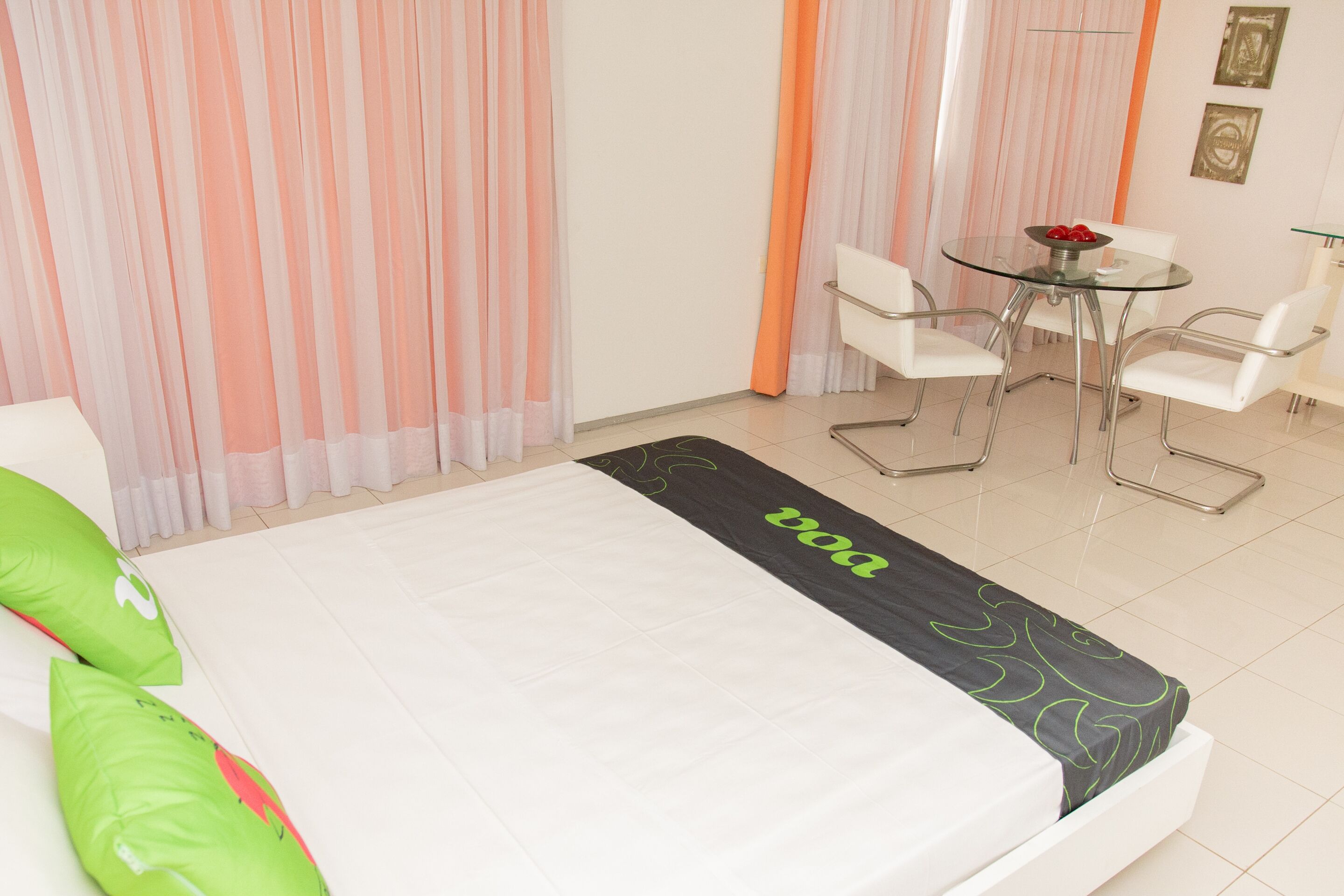 Quarto de hotel com cama, uma mesinha com cadeiras e vista para o exterior através de cortinas finas.