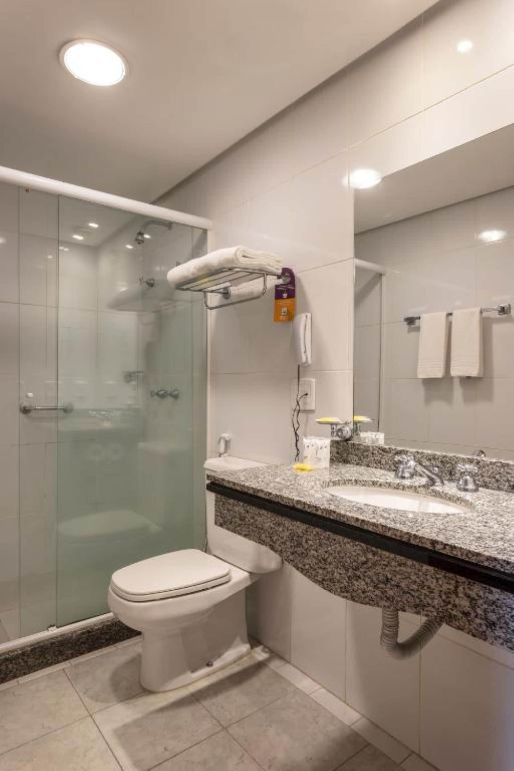 Um banheiro moderno com box de vidro, vaso sanitário branco, bancada de granito e espelho.