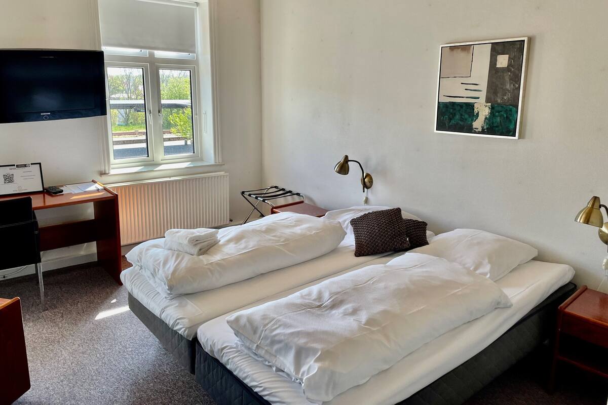 Quarto de hotel com duas camas, uma escrivaninha com computador, televisão e janela com vista para áreas verdes.