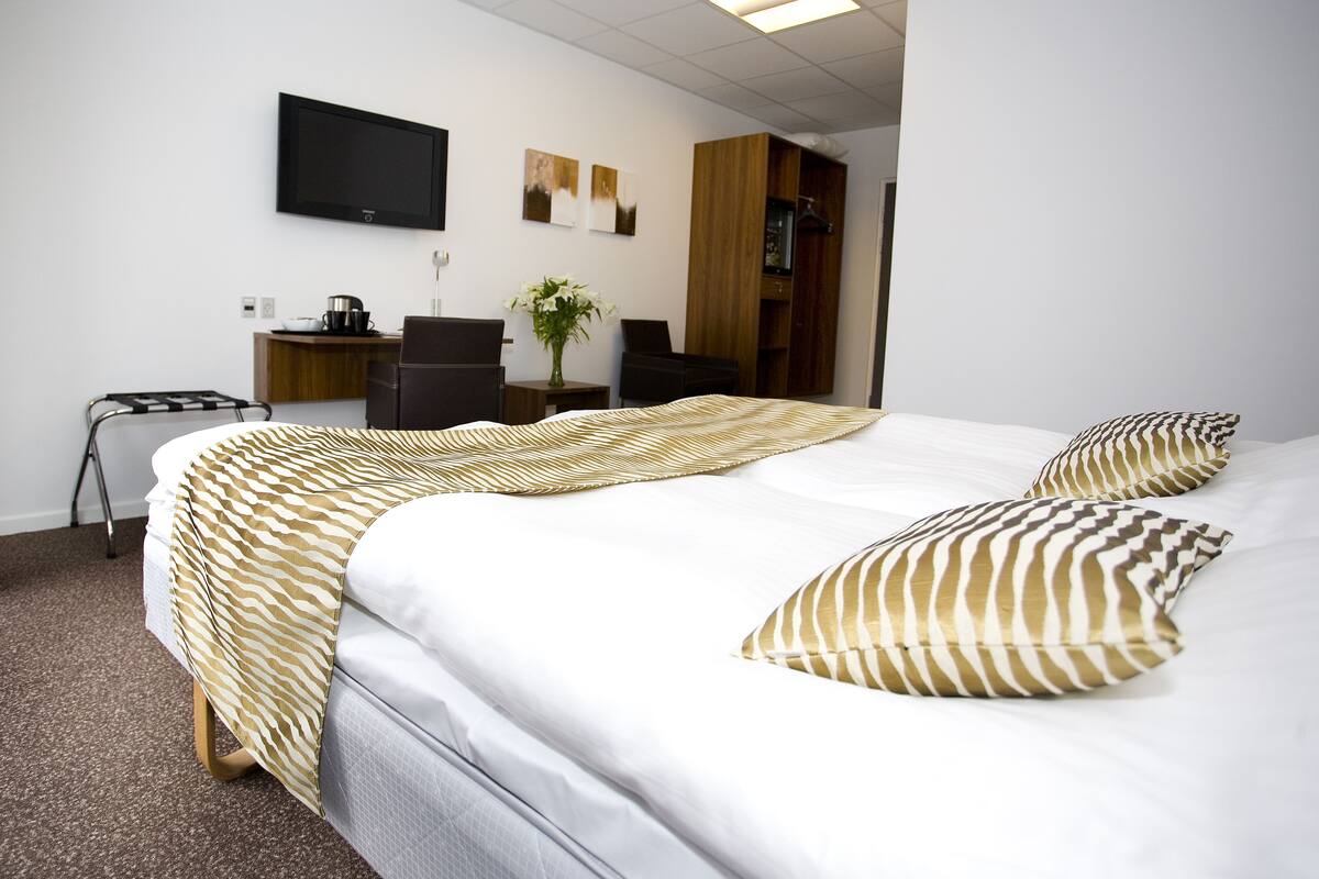 Quarto de hotel com cama, escrivaninha, cadeira, televisão e guarda-roupa.