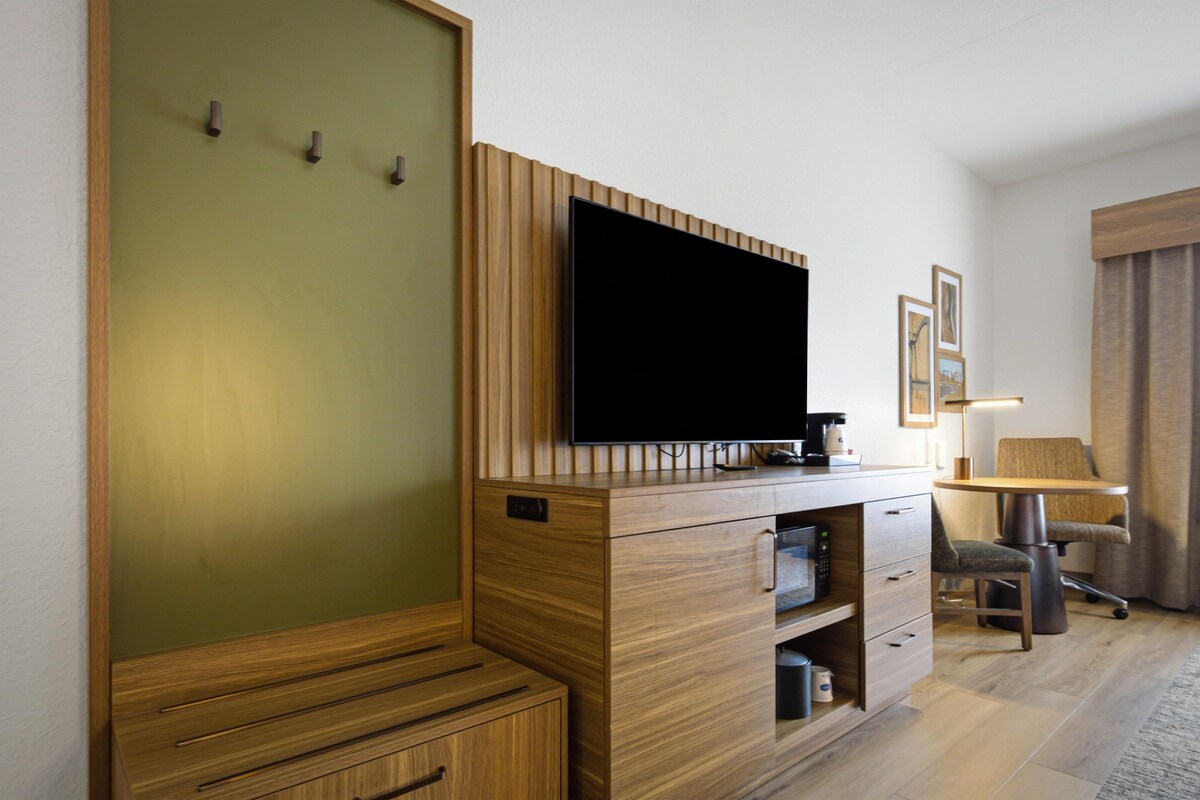 Quarto de hotel com televisão de tela plana fixada em um armário de madeira, uma escrivaninha com luminária, uma cadeira e uma cama com cabeceira de madeira.
