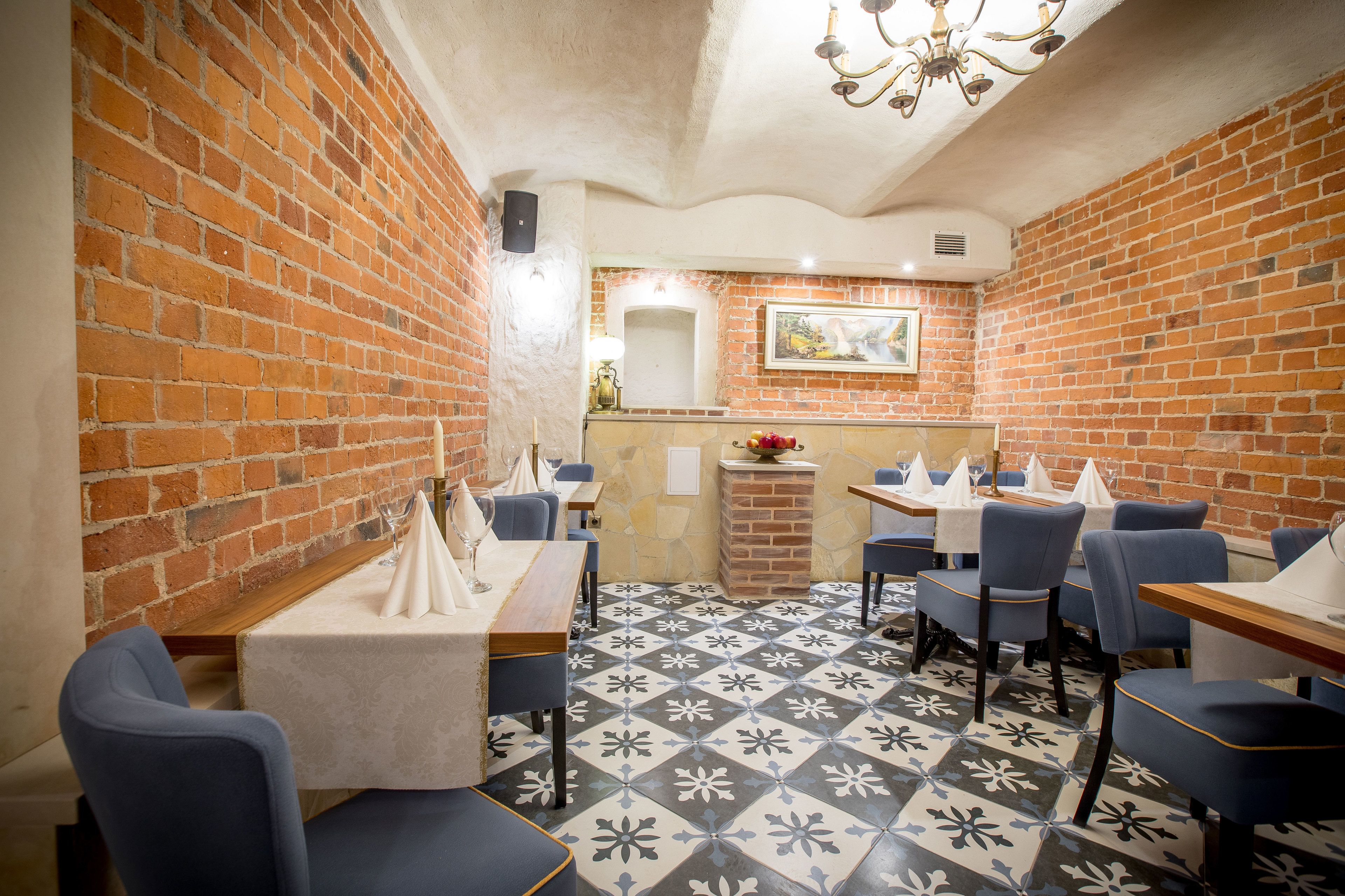 Restaurante com paredes de tijolo, cadeiras azuis e ladrilhos estampados. Mesas montadas com toalhas e guardanapos brancos.