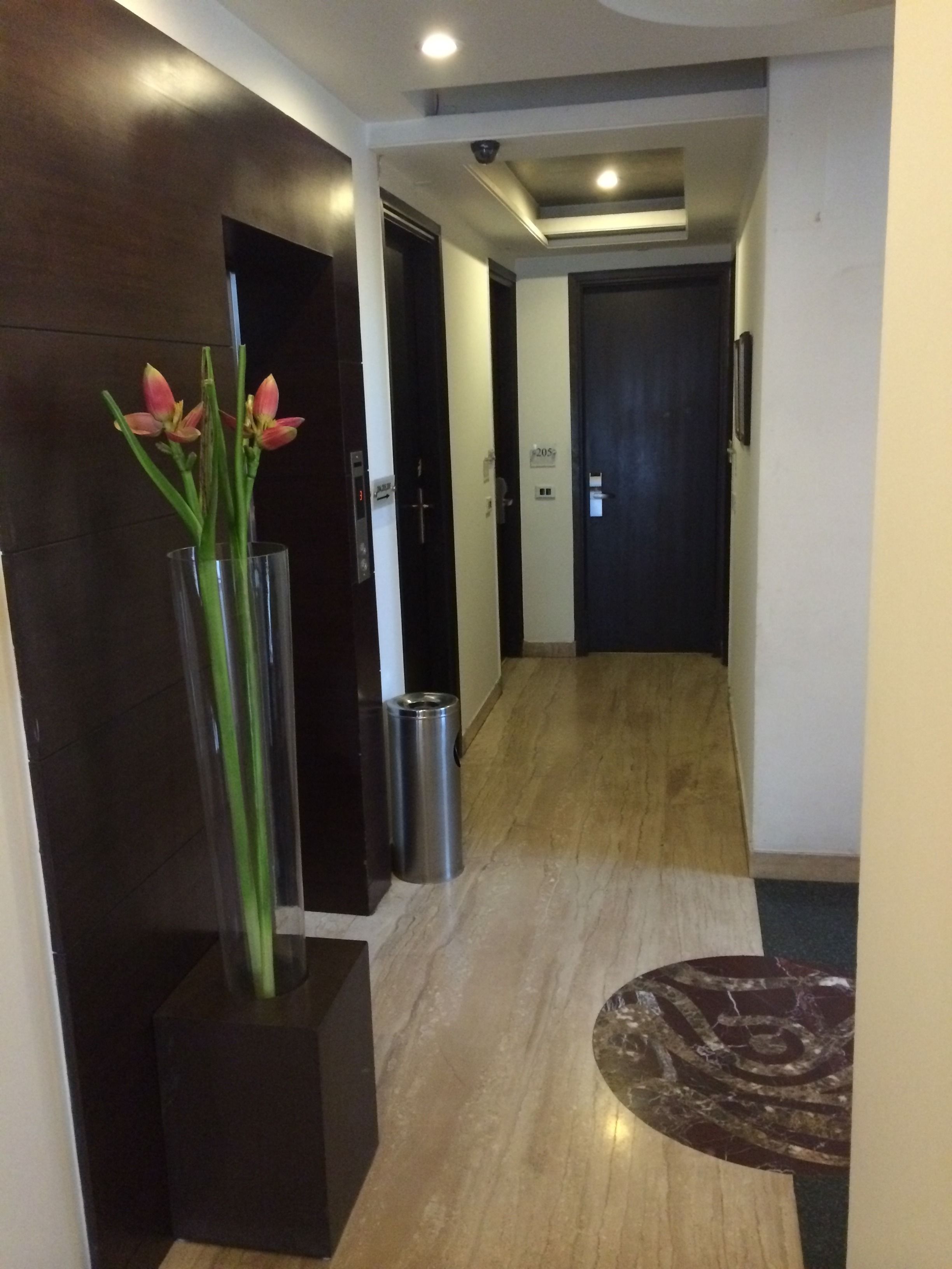 Um corredor com uma porta de madeira escura, um vaso de vidro com flores, uma lixeira e piso de mármore.