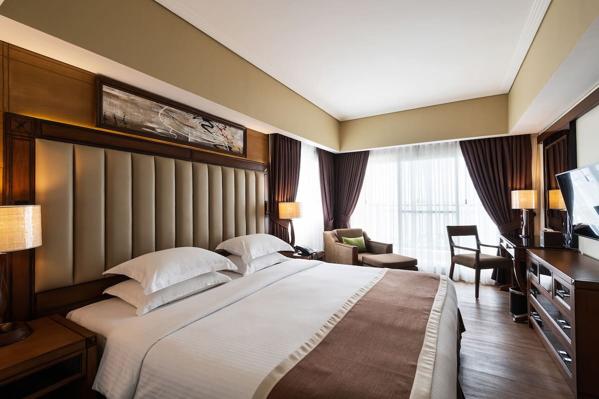 Quarto de hotel com uma cama grande, uma escrivaninha, uma cadeira, uma televisão e uma janela com cortinas.