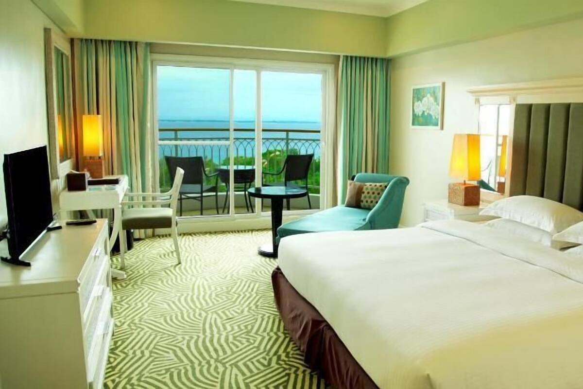 Quarto de hotel com uma cama, escrivaninha, cadeira, televisão e varanda com vista.