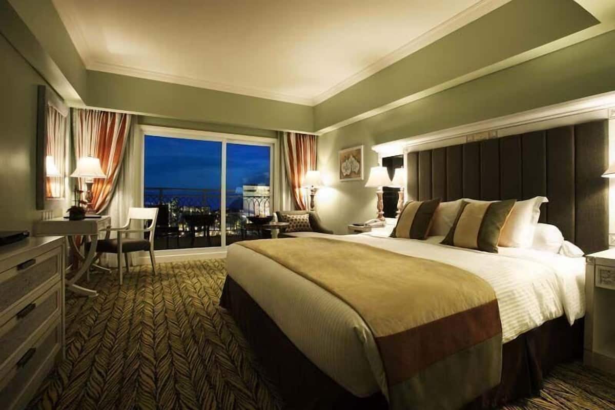 Quarto de hotel com uma cama grande, uma escrivaninha, uma cadeira e vista para a cidade à noite.