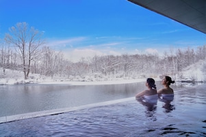 Um casal em piscina aquecida ao ar livre, com paisagem nevada e árvores ao fundo.