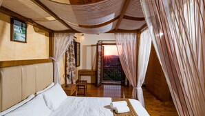Quarto com cama, escrivaninha de madeira, um banquinho e vista para o pôr do sol através de uma porta de varanda.