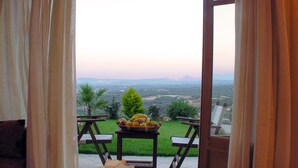 Quarto com vista para uma paisagem verde através de uma abertura. Há uma mesa com uma cesta de frutas e um vaso de flores.