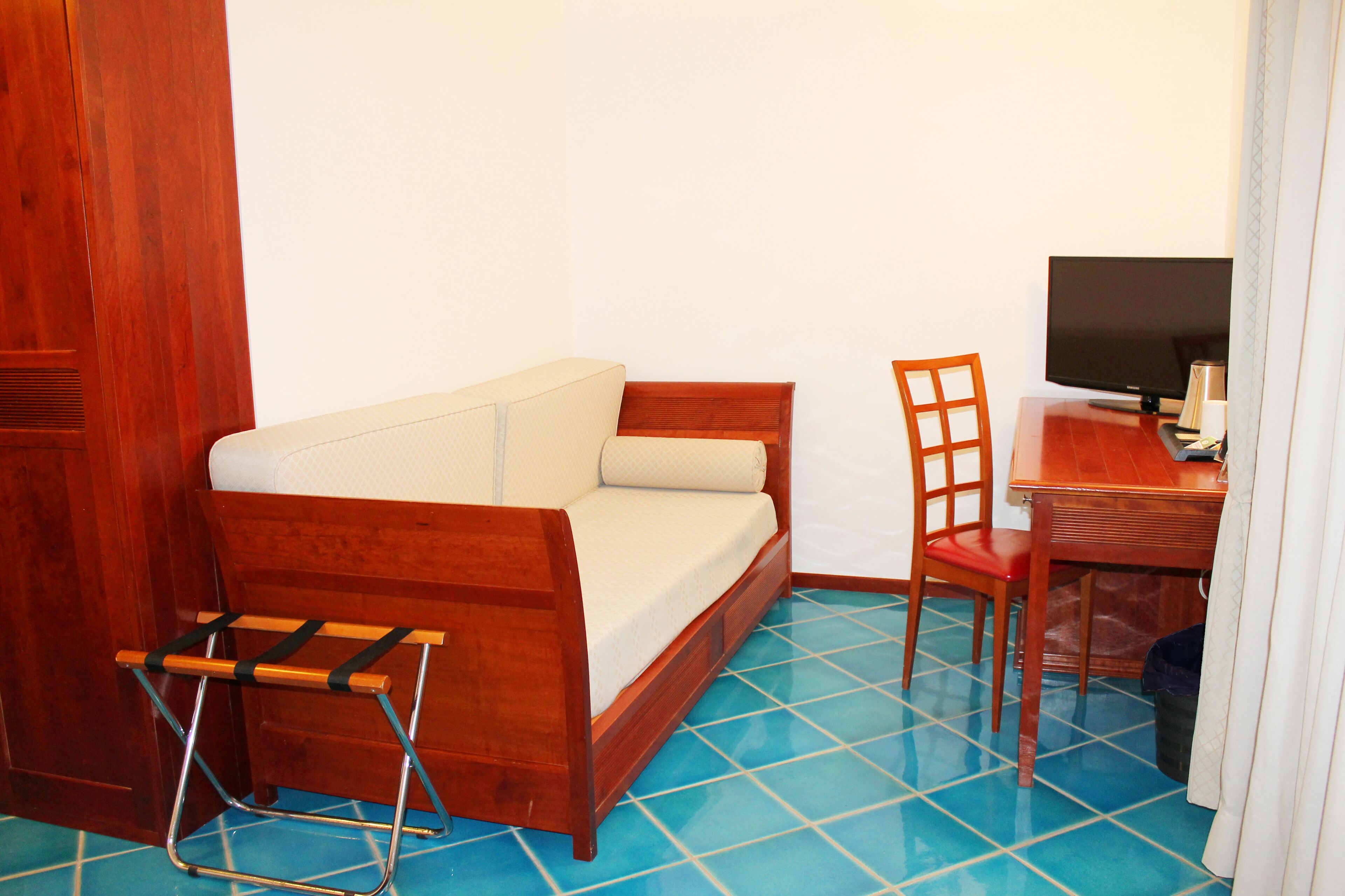 Quarto com sofá de madeira, cadeira de madeira, mesa pequena com televisão e chão de azulejos azuis.
