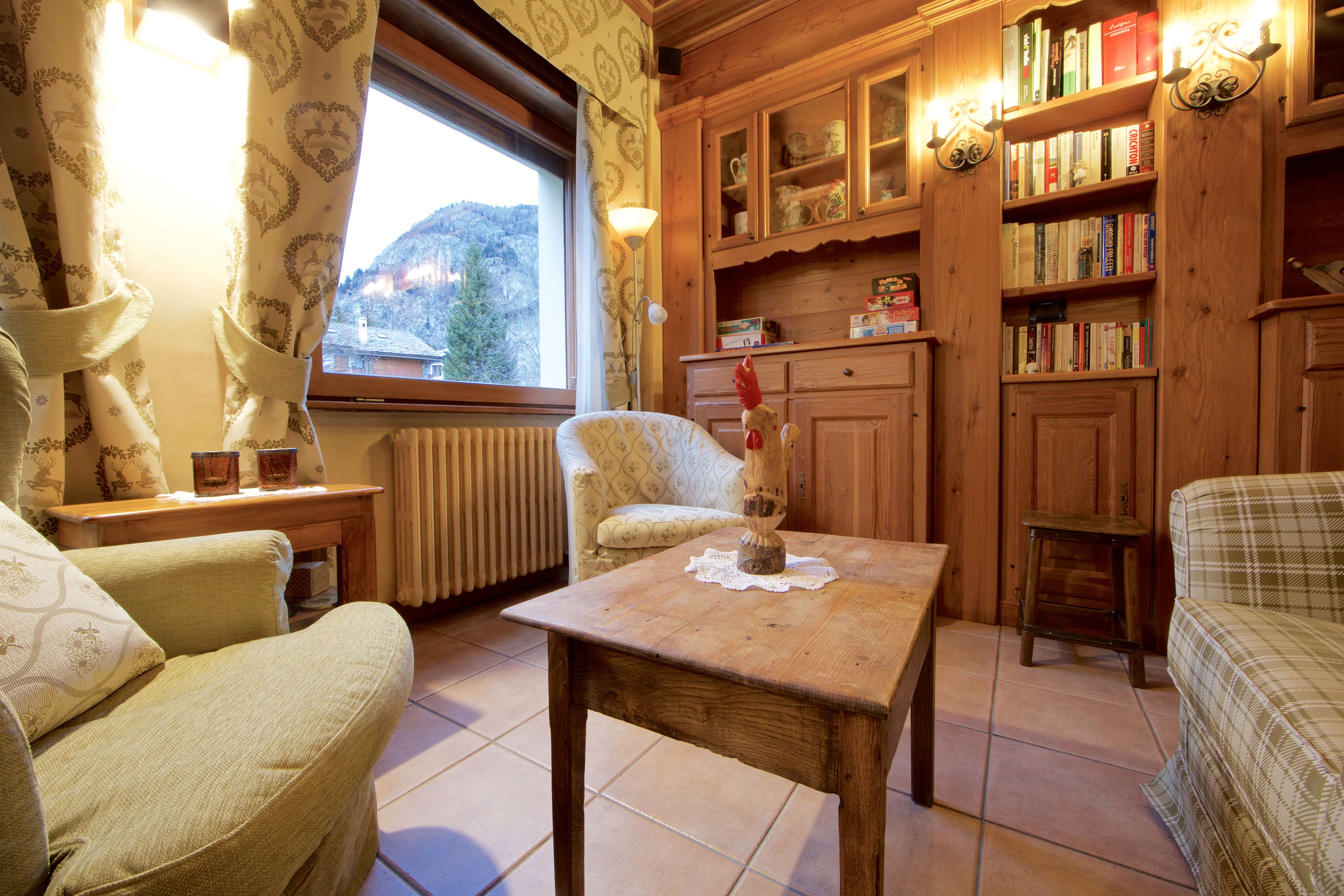 Uma sala de estar acolhedora com uma mesa de madeira, duas poltronas, uma estante de livros e vista para uma montanha pela janela.