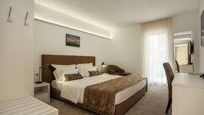 Quarto de hotel com uma cama grande, uma escrivaninha, uma cadeira, uma mesinha de cabeceira e uma luminária.
