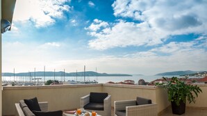 Um terraço com mobiliário de vime, uma mesa com itens de café da manhã e vista para um porto e colinas ao longe.
