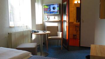 Quarto de hotel com uma cama, uma mesinha com uma cadeira, uma televisão presa à parede e um banheiro visível por uma porta aberta.