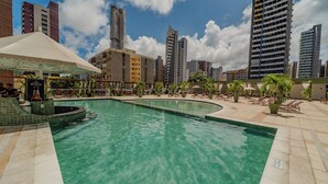 Uma piscina no terraço com toldo branco, cercada por edifícios altos e palmeiras.