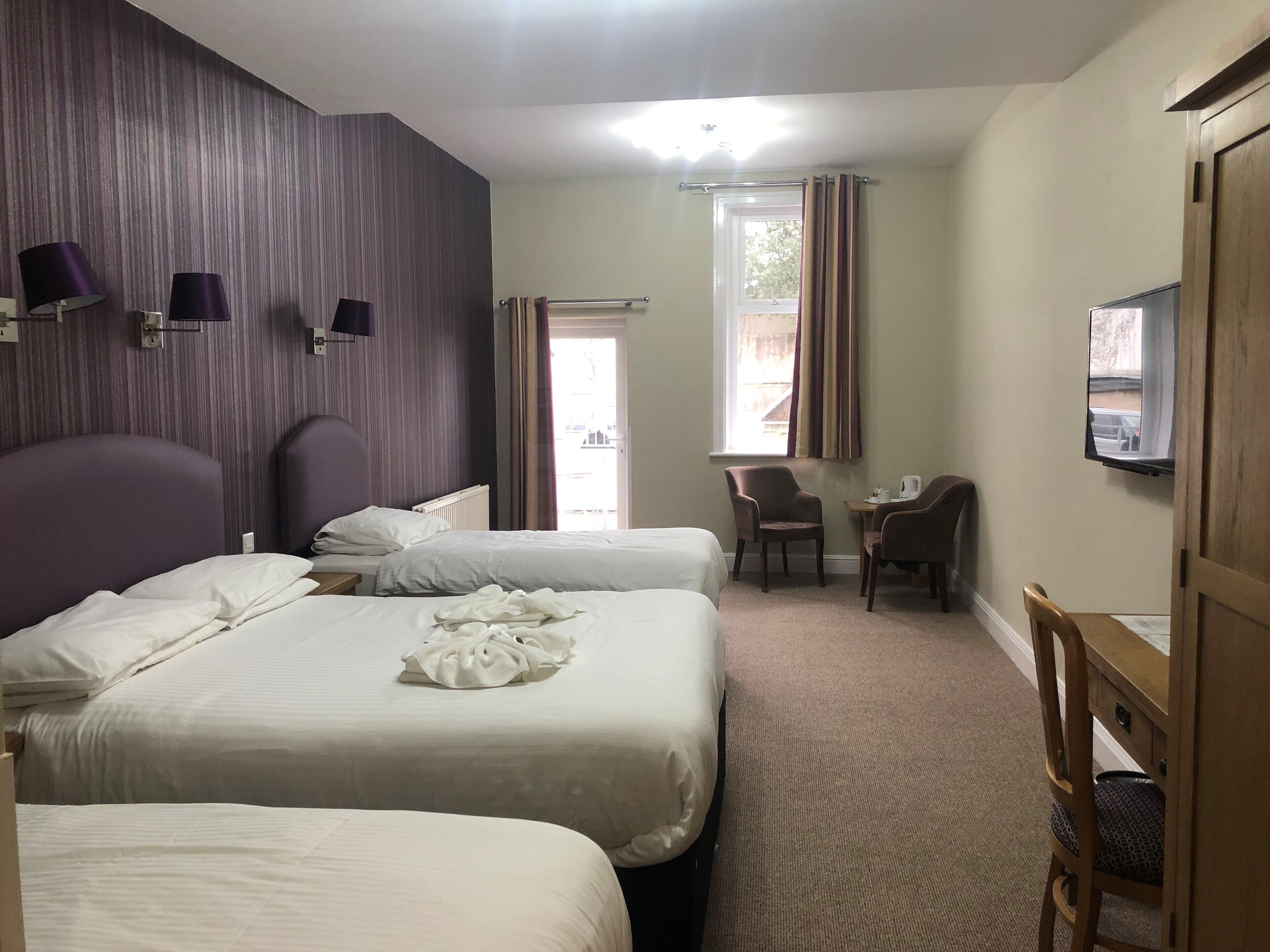 Quarto de hotel com duas camas, uma escrivaninha e uma cadeira. Há uma janela com cortinas, um radiador e uma TV de tela plana na parede.