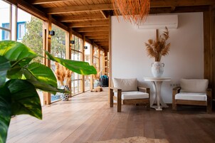 Quarto com piso de madeira, uma mesa branca com um vaso de capim-do-campo, duas cadeiras brancas acolchoadas e grandes janelas que permitem a entrada de luz natural.