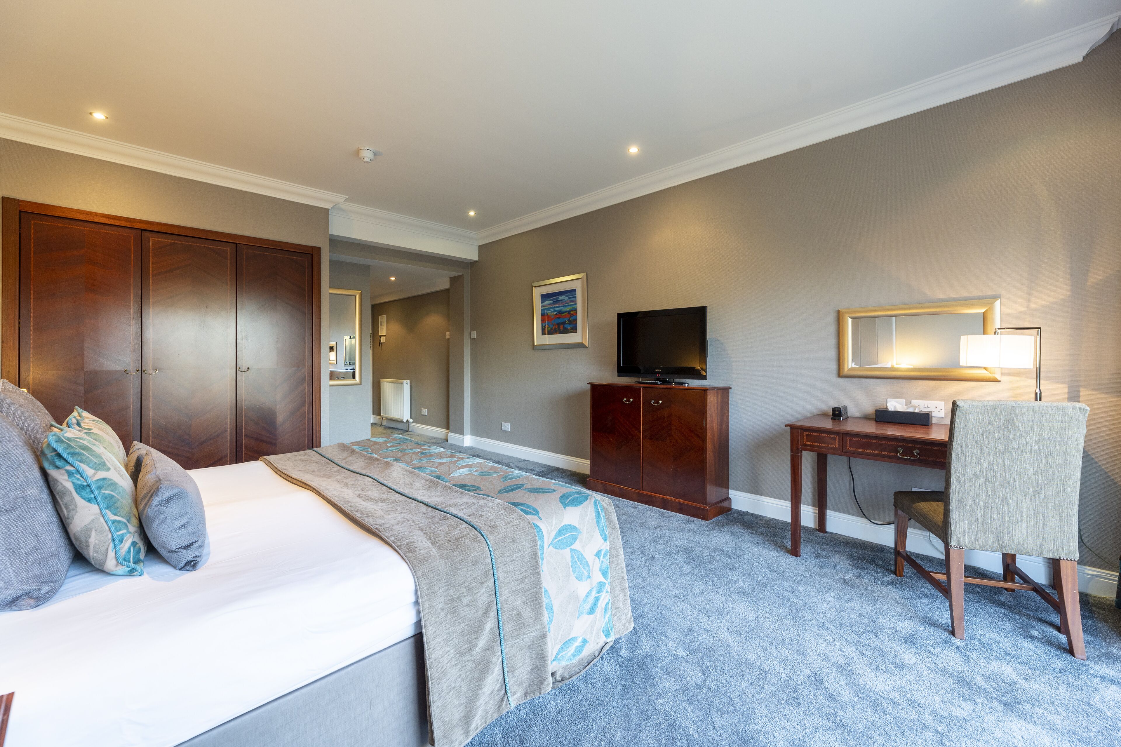 Quarto de hotel com cama, escrivaninha, cadeira, guarda-roupa, televisão e um quadro na parede.
