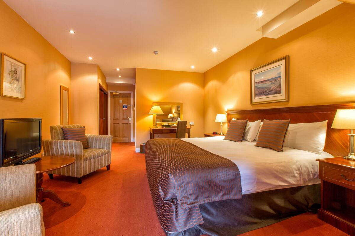 Quarto de hotel com cama, duas poltronas, televisão, escrivaninha e abajur.