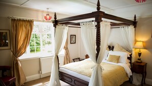 Uma cama com dossel sobre pilares, estrutura de madeira, roupa de cama branca e mesa de cabeceira com abajur.