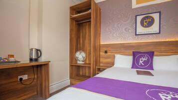 Quarto de hotel com cama, escrivaninha de madeira, chaleira, ventilador e parede com um logotipo.
