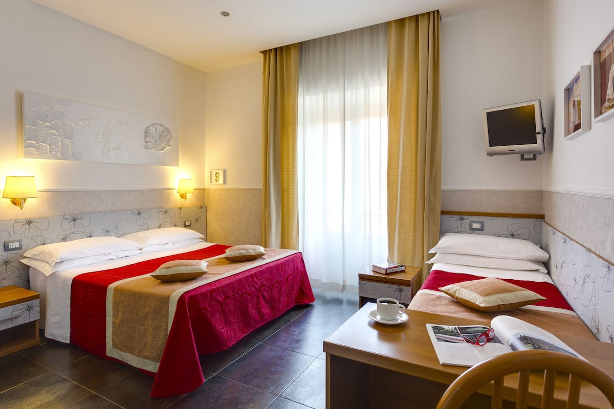 Quarto de hotel com duas camas, uma escrivaninha, uma cadeira, uma televisão e uma janela com cortinas.