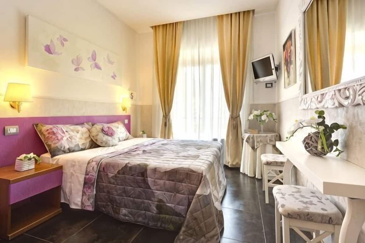 Quarto de hotel com cama, mesa de cabeceira, luminária, escrivaninha, cadeira e televisão.