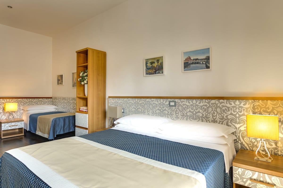Quarto de hotel com duas camas, cabeceira de madeira, mesa de cabeceira com abajur e dois quadros na parede.