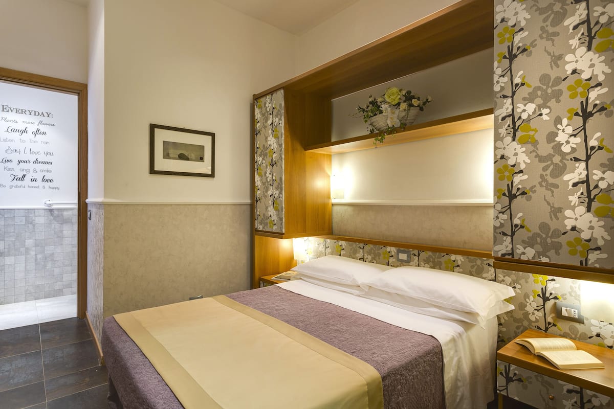 Cama bem arrumada com colcha roxa e bege, mesa de cabeceira com um livro, e parede com papel de parede estampado com flores.