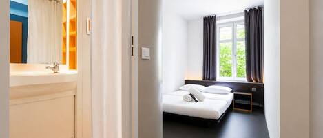 Quarto de hotel com cama, janela, mesa de cabeceira e banheiro com pia e espelho.