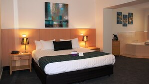 Quarto de hotel com uma cama grande, duas luminárias de cabeceira, uma mesinha de cabeceira, um aparador de madeira e banheiro com banheira e pia.