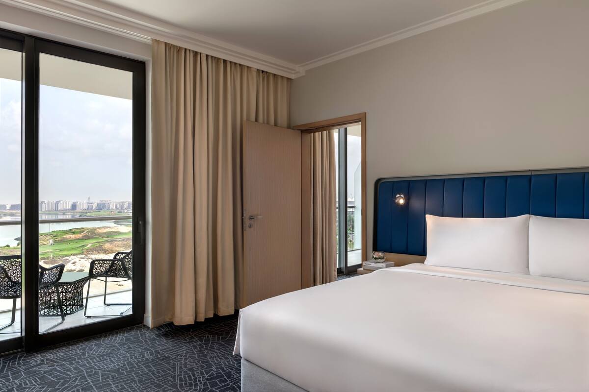 Quarto de hotel com uma cama grande, vista para a paisagem urbana através de uma porta de correr de vidro e varanda com mobiliário de exterior.