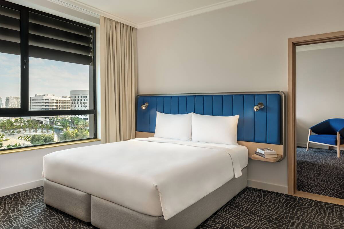 Quarto de hotel com uma cama grande, vista para a cidade pela janela, cabeceira azul e um espelho na parede.
