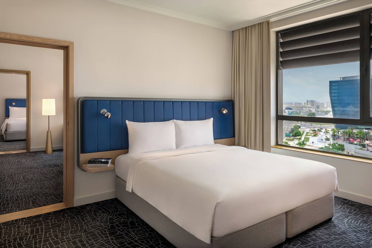 Quarto de hotel com uma cama grande, uma mesa de cabeceira, um espelho, uma janela com vista para a cidade e uma porta.