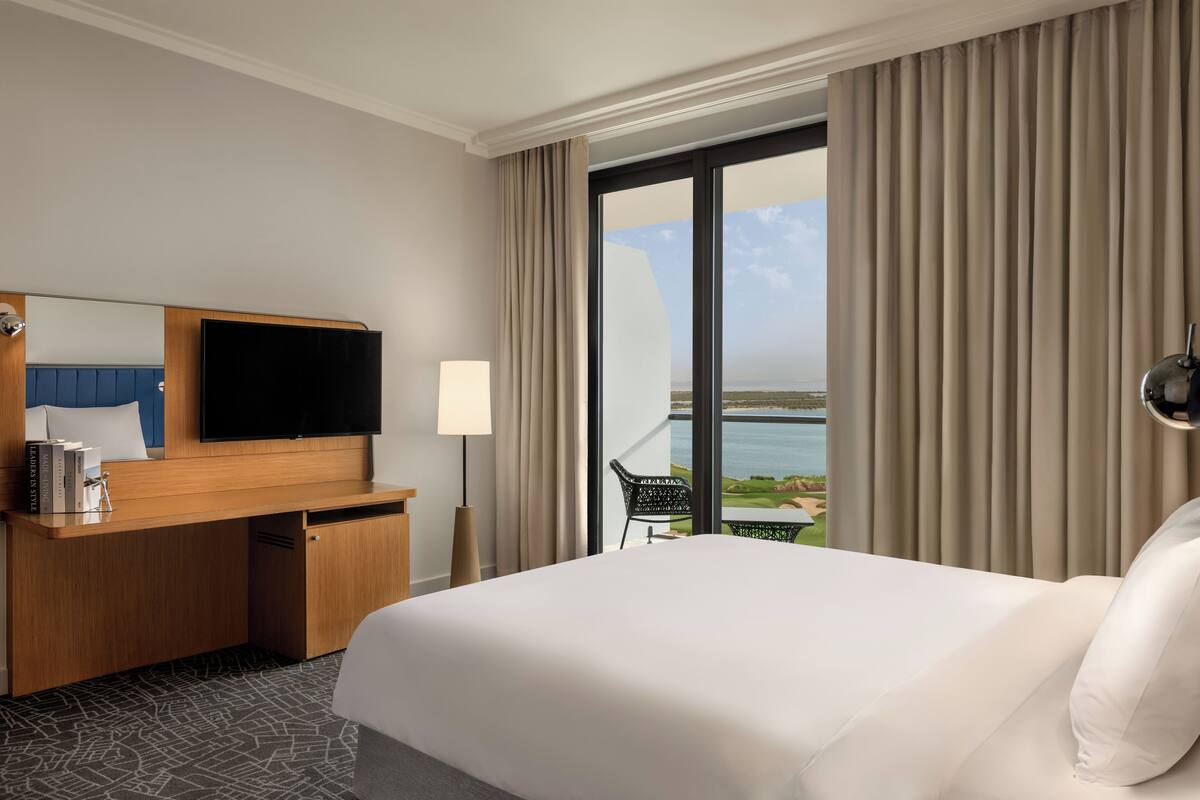 Quarto de hotel com uma cama grande, uma escrivaninha com televisão de tela plana, uma cadeira e vista para o mar por janelas amplas.