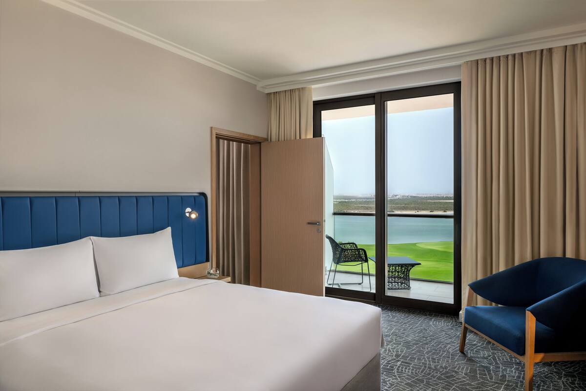 Quarto de hotel com uma cama grande, cabeceira azul, porta de madeira e vista para um campo de golfe através de uma porta de vidro deslizante.