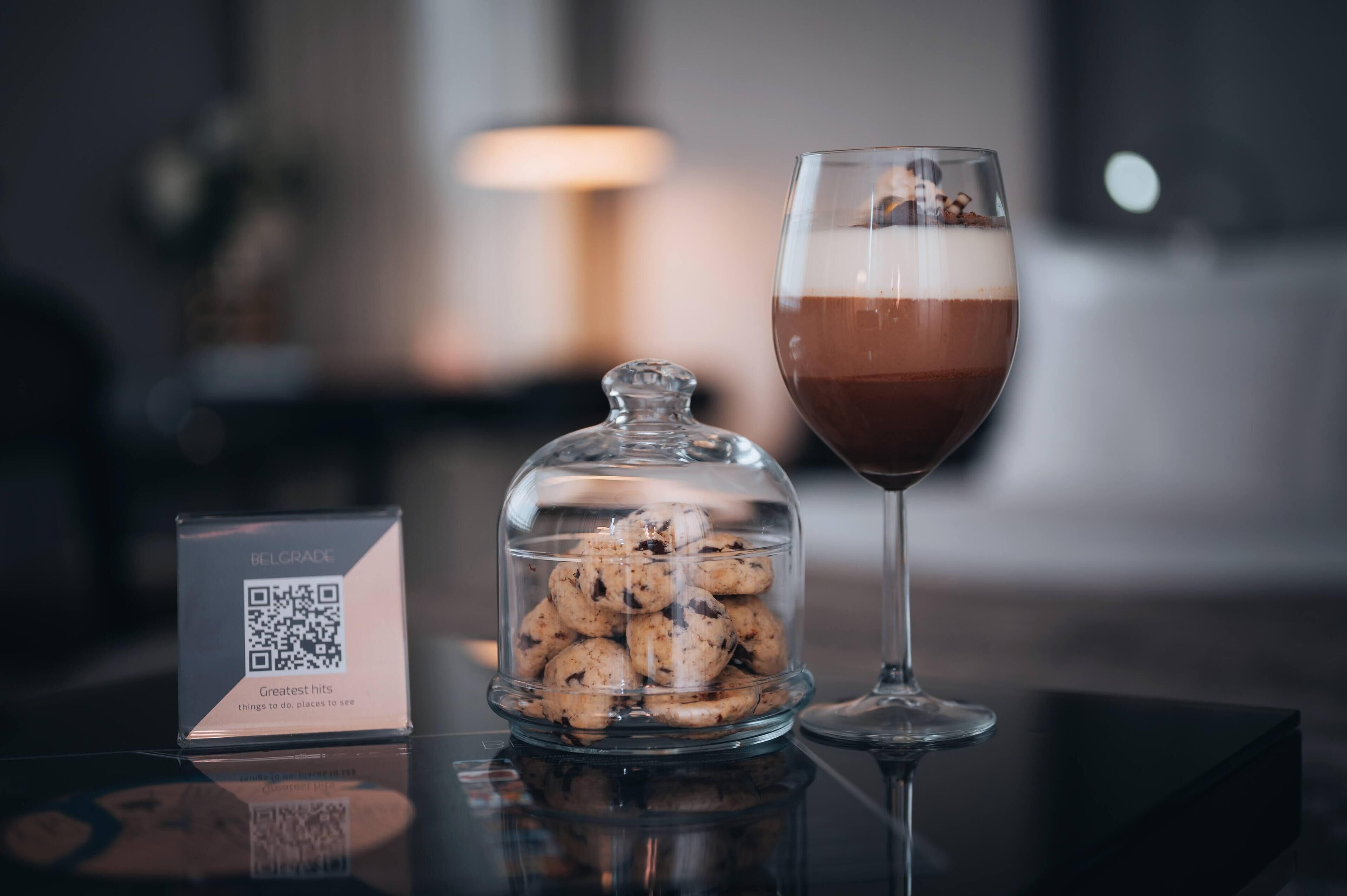 Um copo com bebida em camadas, um pote de biscoitos e uma placa com código QR em uma superfície reflexiva.
