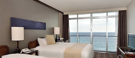Quarto de hotel com duas camas, uma janela grande com vista para o mar, uma escrivaninha com um computador e uma televisão.