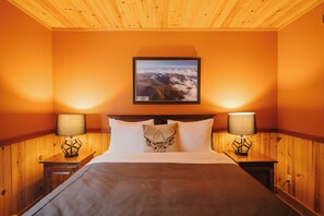 Quarto com teto de madeira, cama com roupa de cama branca, duas mesas de cabeceira com abajures e um quadro na parede.