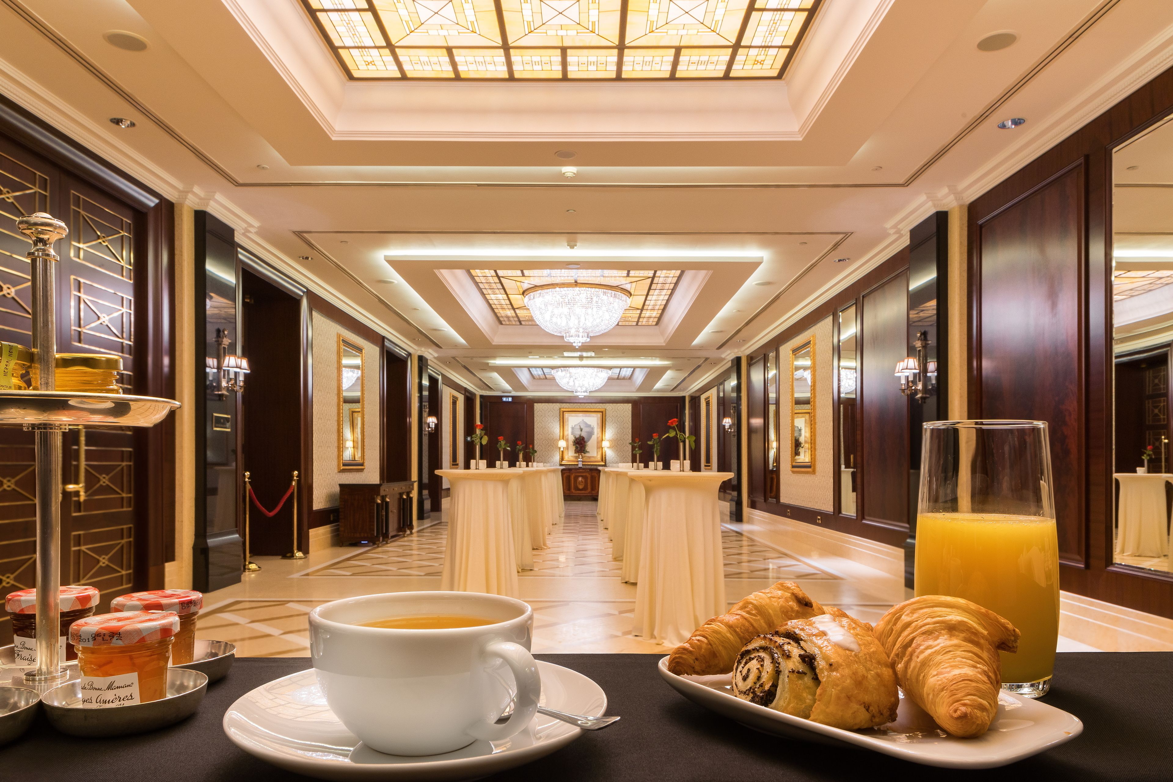 Um saguão de hotel com um corredor comprido, um candelabro e uma mesa de pequeno-almoço com croissants e sumo de laranja.