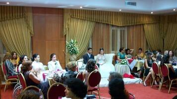 Um evento formal em um salão de festas com mesas redondas, cadeiras vermelhas e um palco com fundo branco.