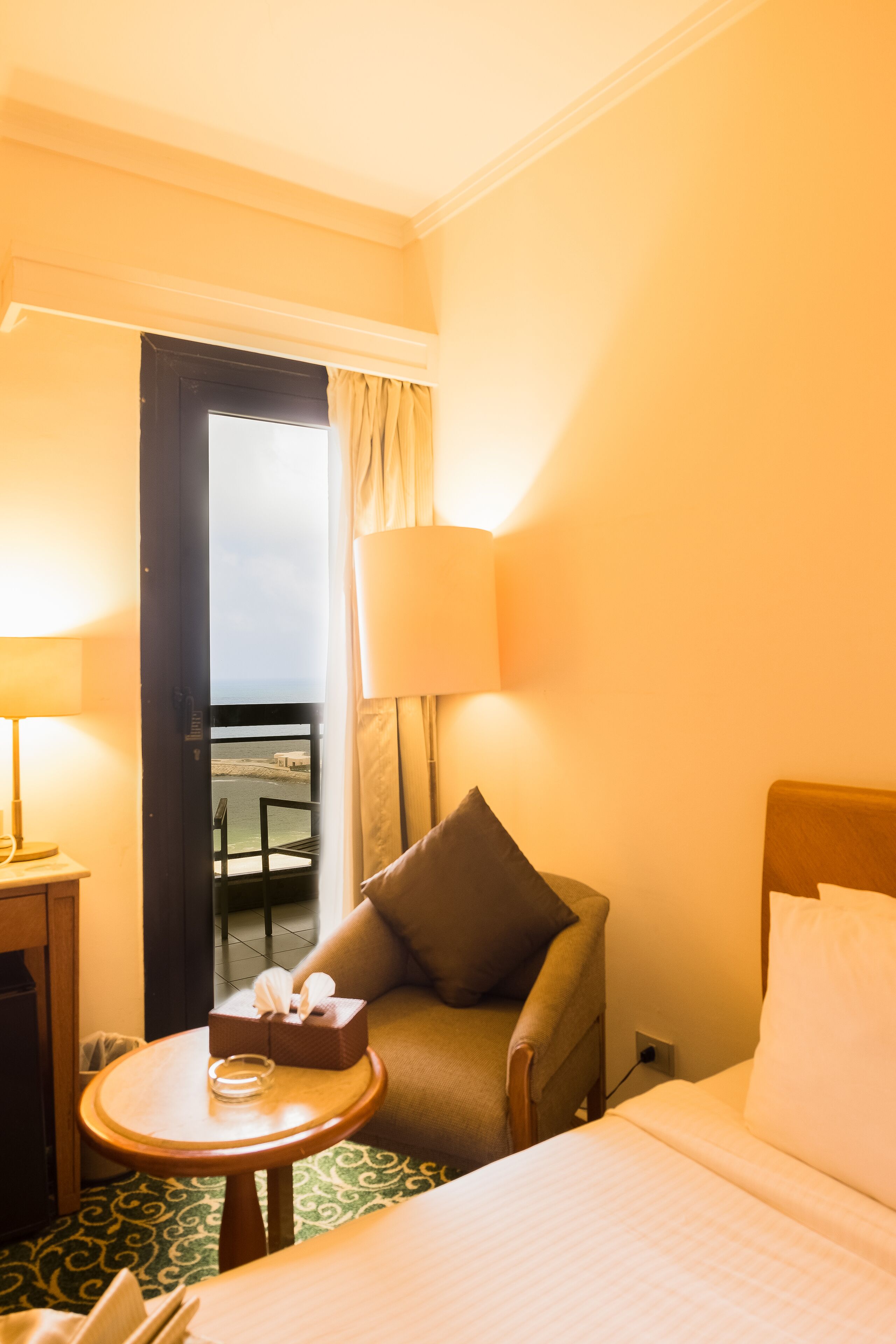 Quarto de hotel com cama, uma cadeira, uma pequena mesa com caixa de lenços e vista para o mar através de uma porta aberta.