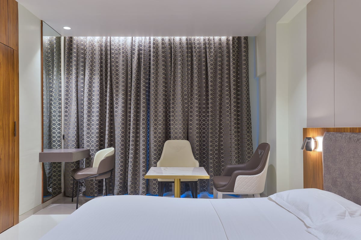 Quarto de hotel com uma cama, uma escrivaninha, duas cadeiras e uma janela grande com cortinas estampadas.